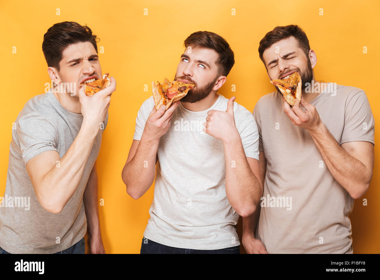 Three young smiling men eating pizza isolated over yellow background ...