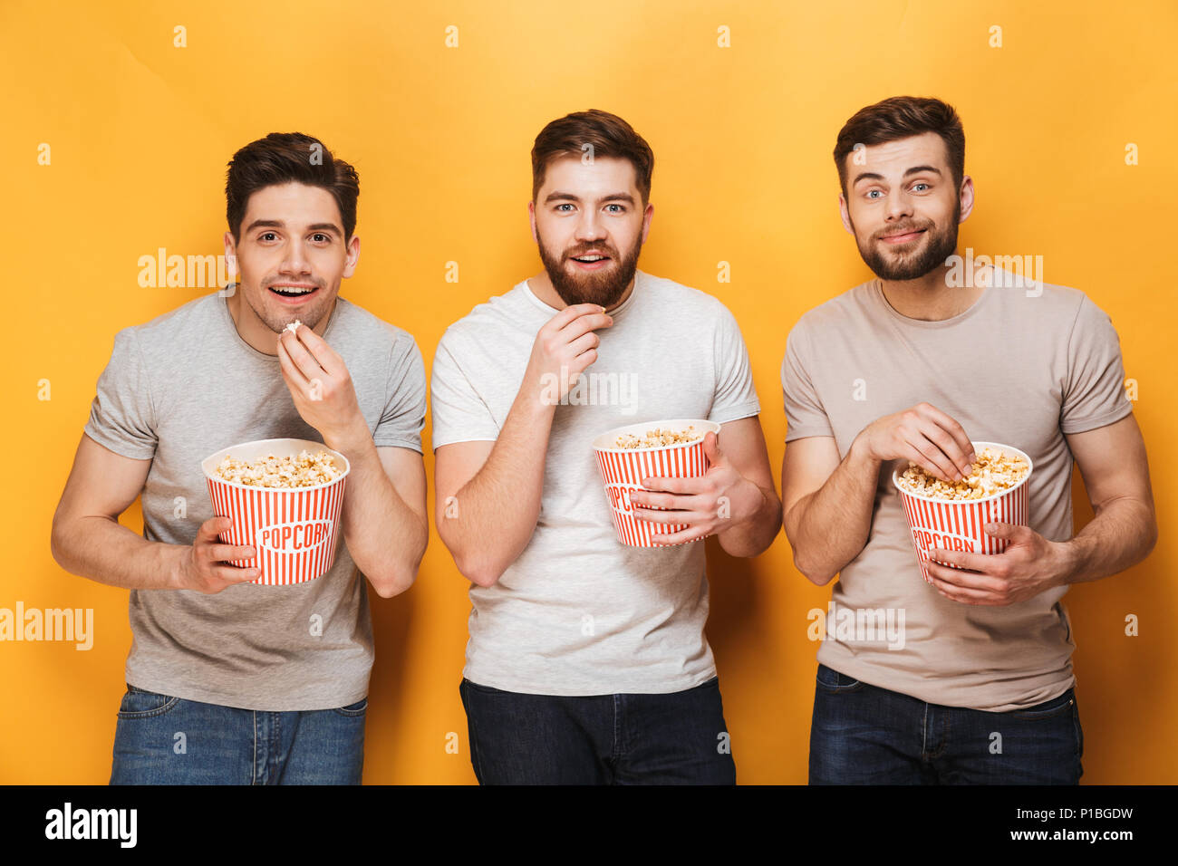 Three young smiling men eating popcorn and looking at camera isolated ...