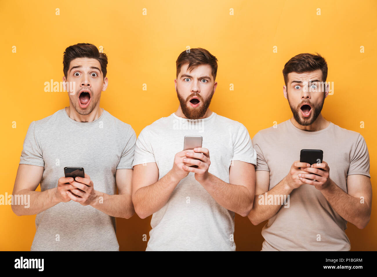 Three young shocked men holding mobile phones isolated over yellow ...