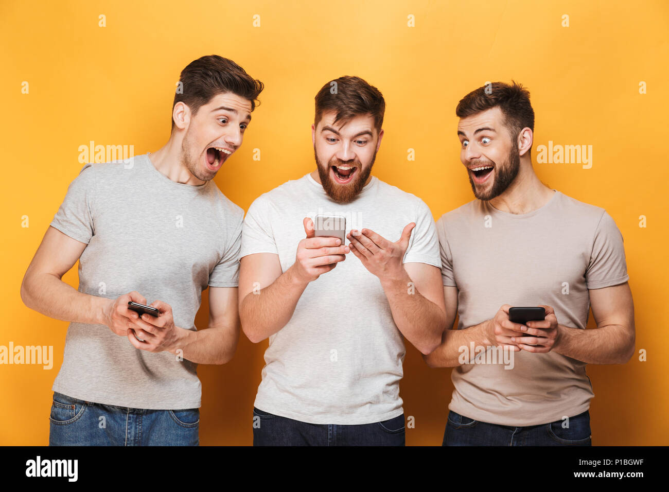 Three young cheerful men holding mobile phones isolated over yellow ...