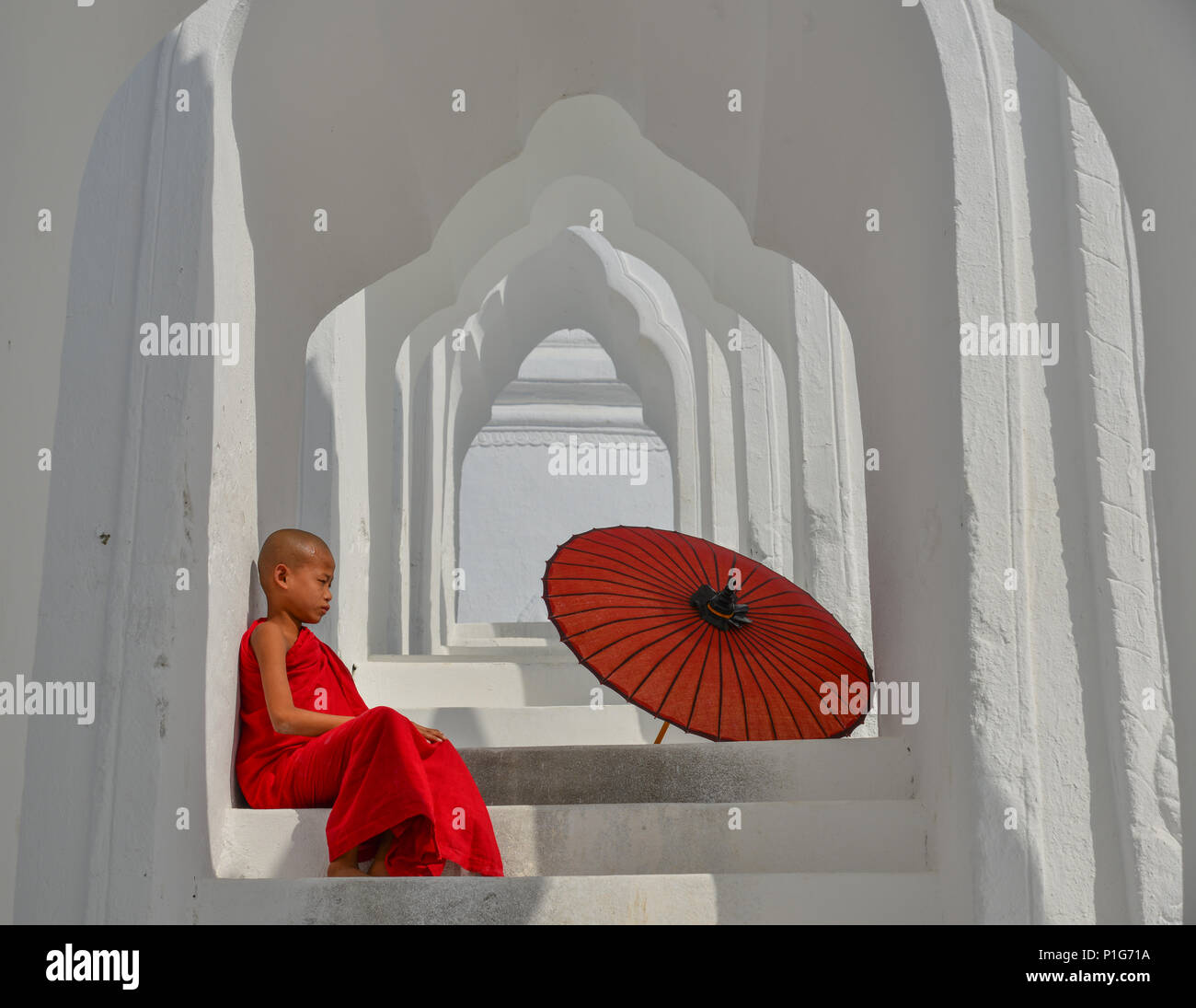 A Burmese Buddhist novice monk with a red umbrella sitting at the white ...