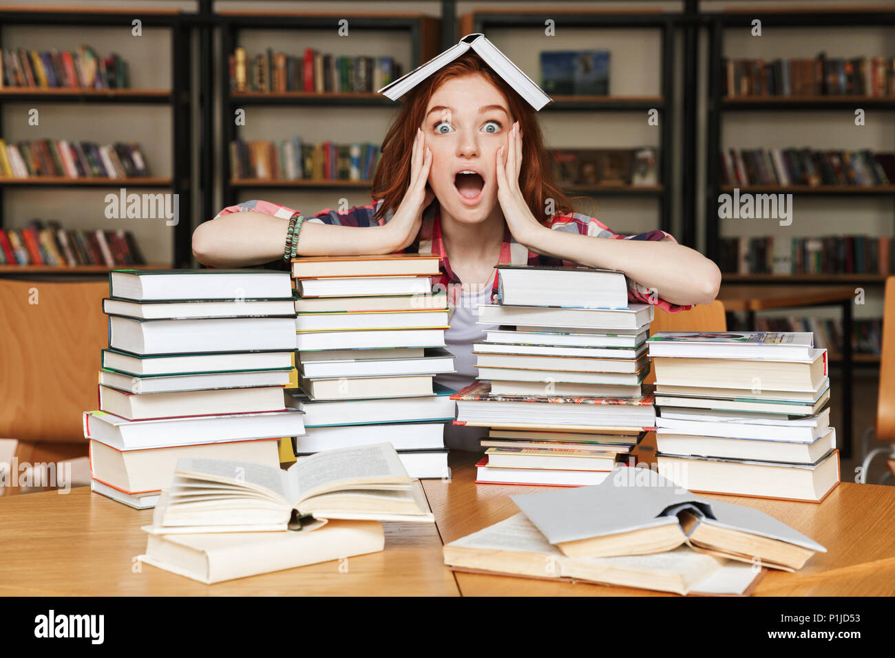 Surprised teenage girl sitting at the library table with big stacks of ...
