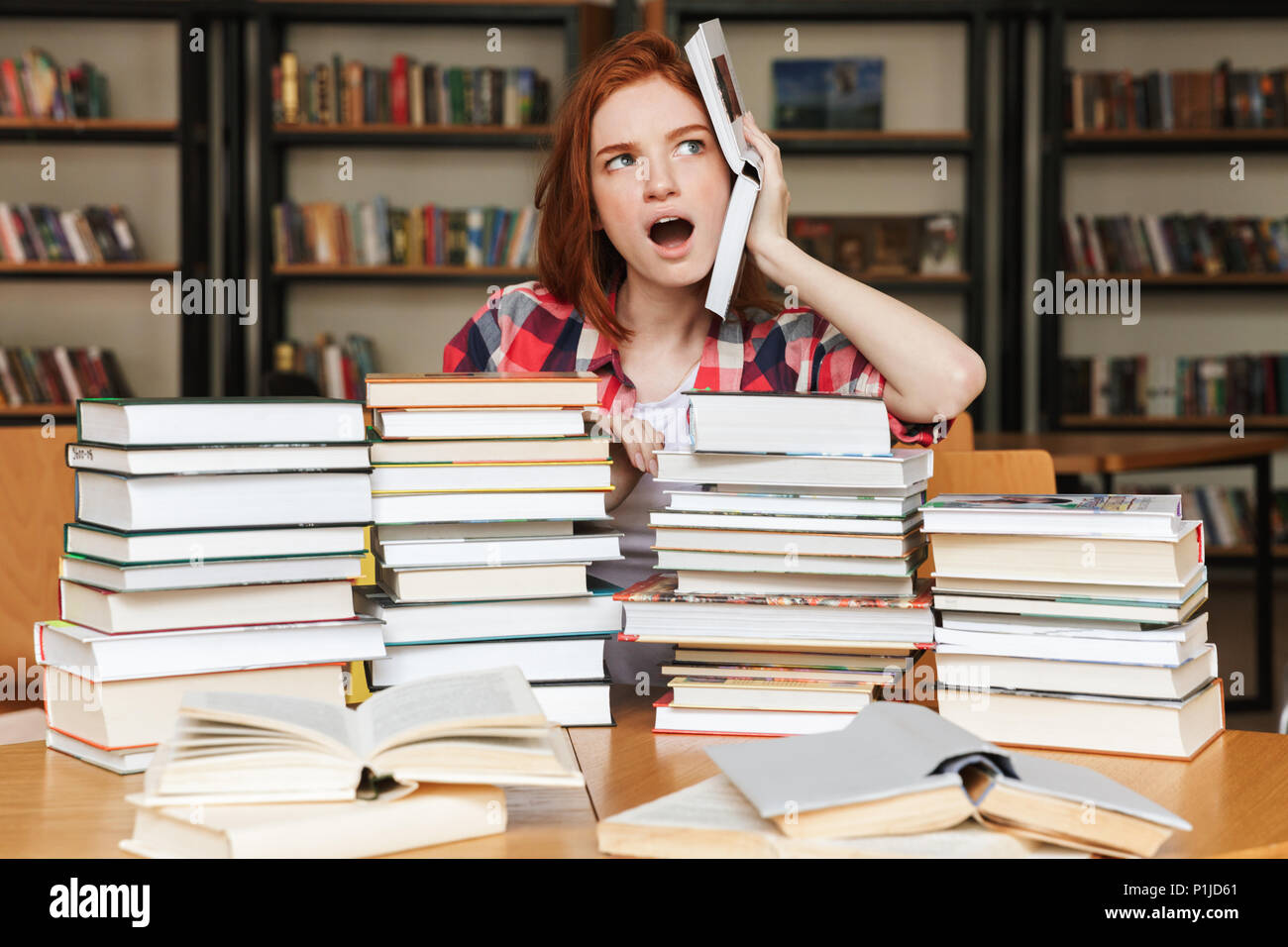 Confused teenage girl sitting at the library table with big stacks of ...