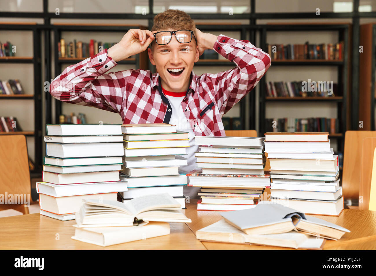 Cheerful teenage boy sitting at the library table with big stacks of ...