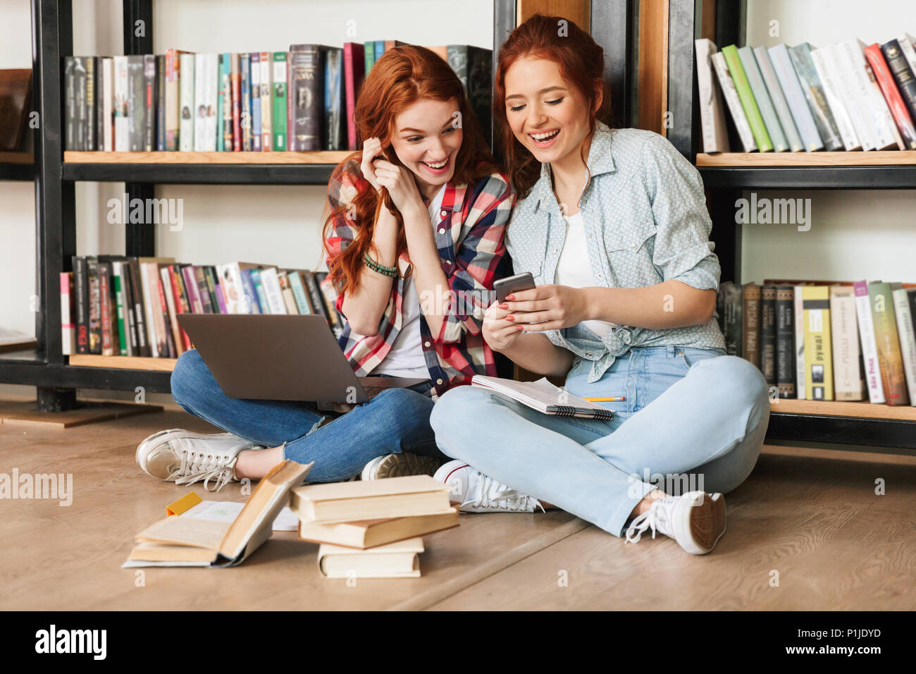 Two laughing teenage girls sitting on a floor at the bookshelf in a ...
