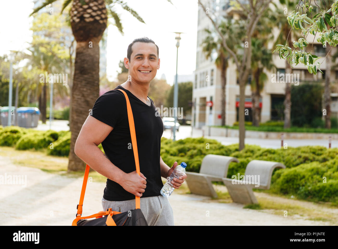Image of cheerful young strong sports man walking outdoors with bottle ...