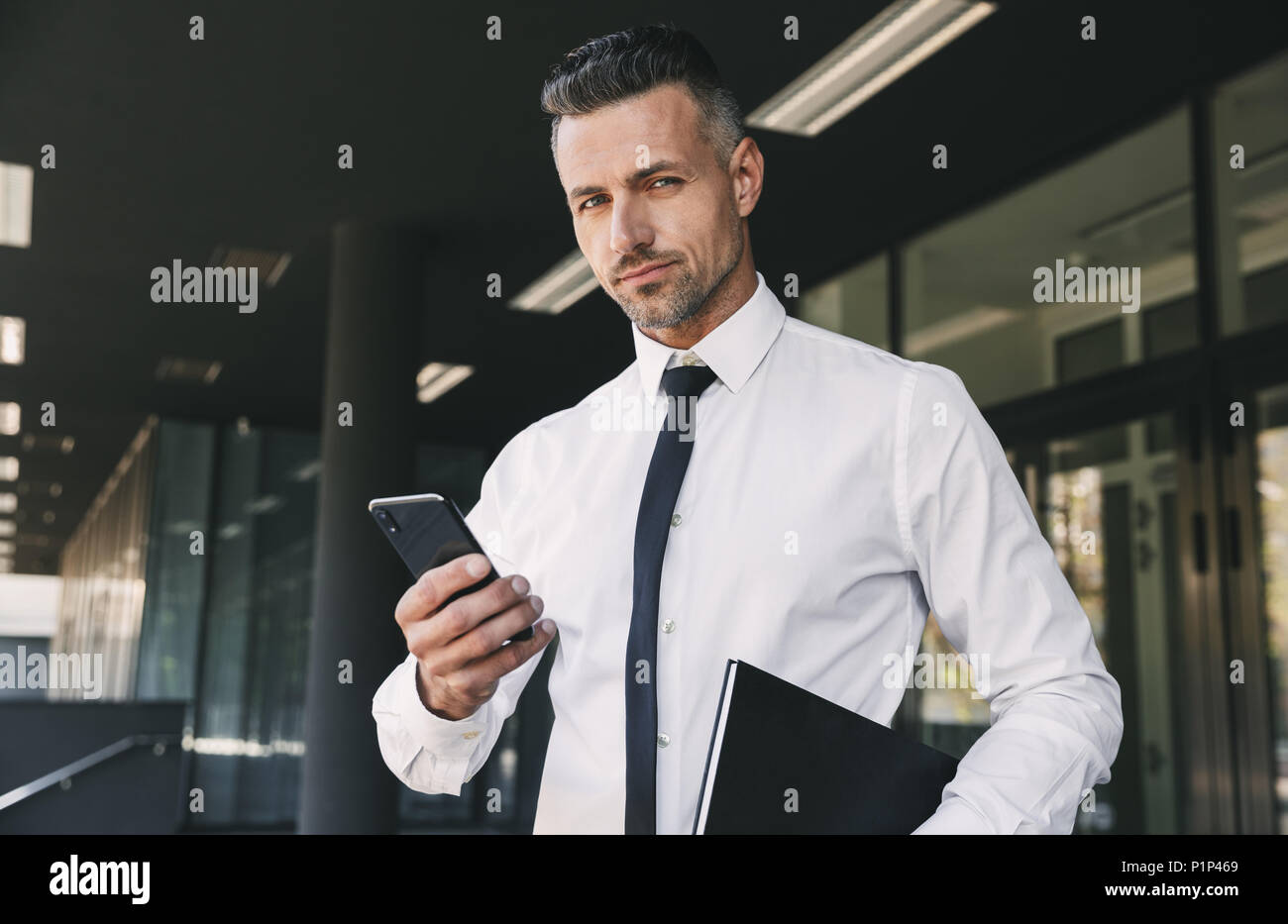 Portrait of a confident young businessman dressed in formal clothes standing outside a glass ...