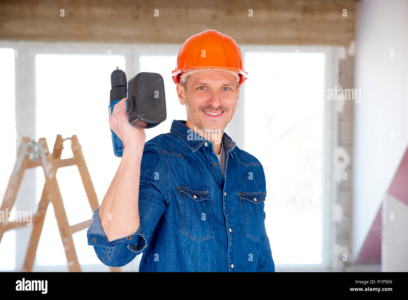 Smiling handyman wearing safety helmet and holding drill in his hand ...
