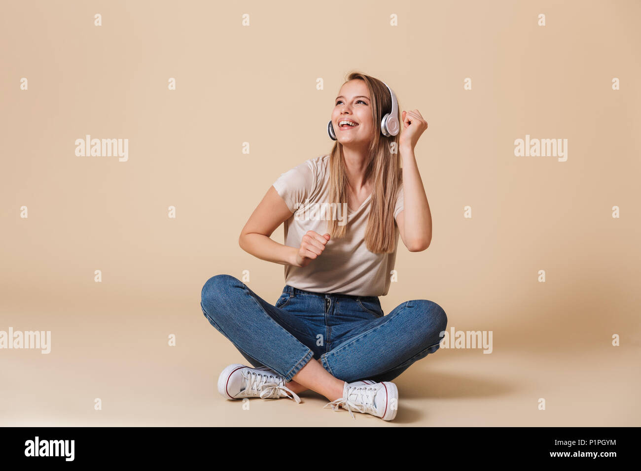 Cheerful woman 20s in jeans singing and dancing while sitting on floor ...
