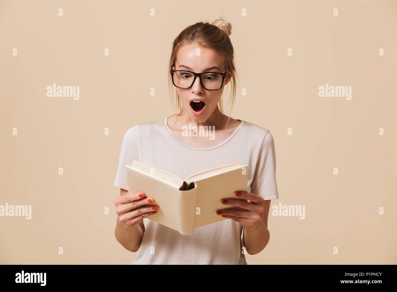 Portrait of a shocked young girl in eyewear reading a book isolated ...