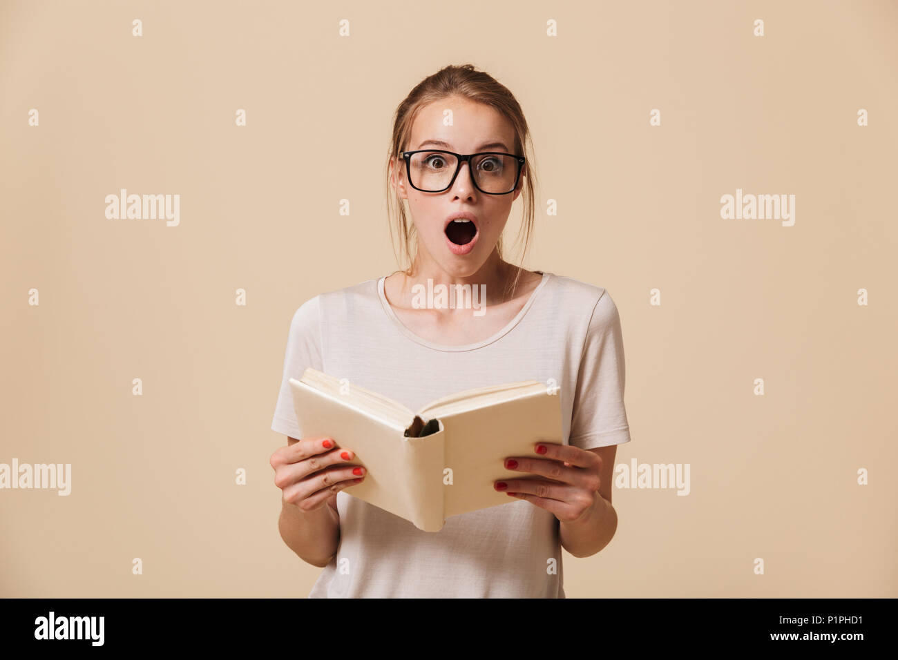 Portrait of a surprised young girl in eyewear reading a book isolated ...
