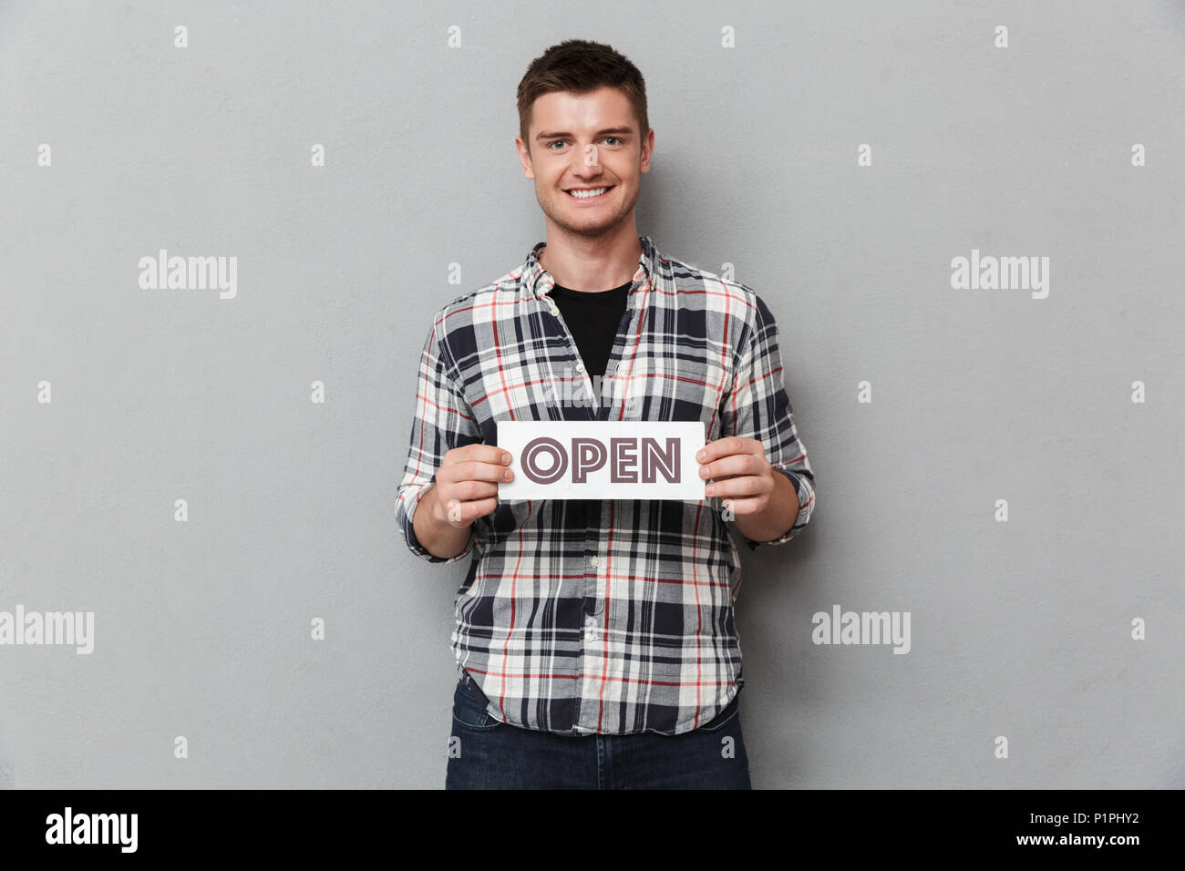 Portrait of a smiling young man showing board with open sign over gray ...