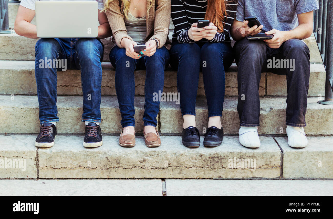 Four students sitting in a row on a step using their technology on the ...