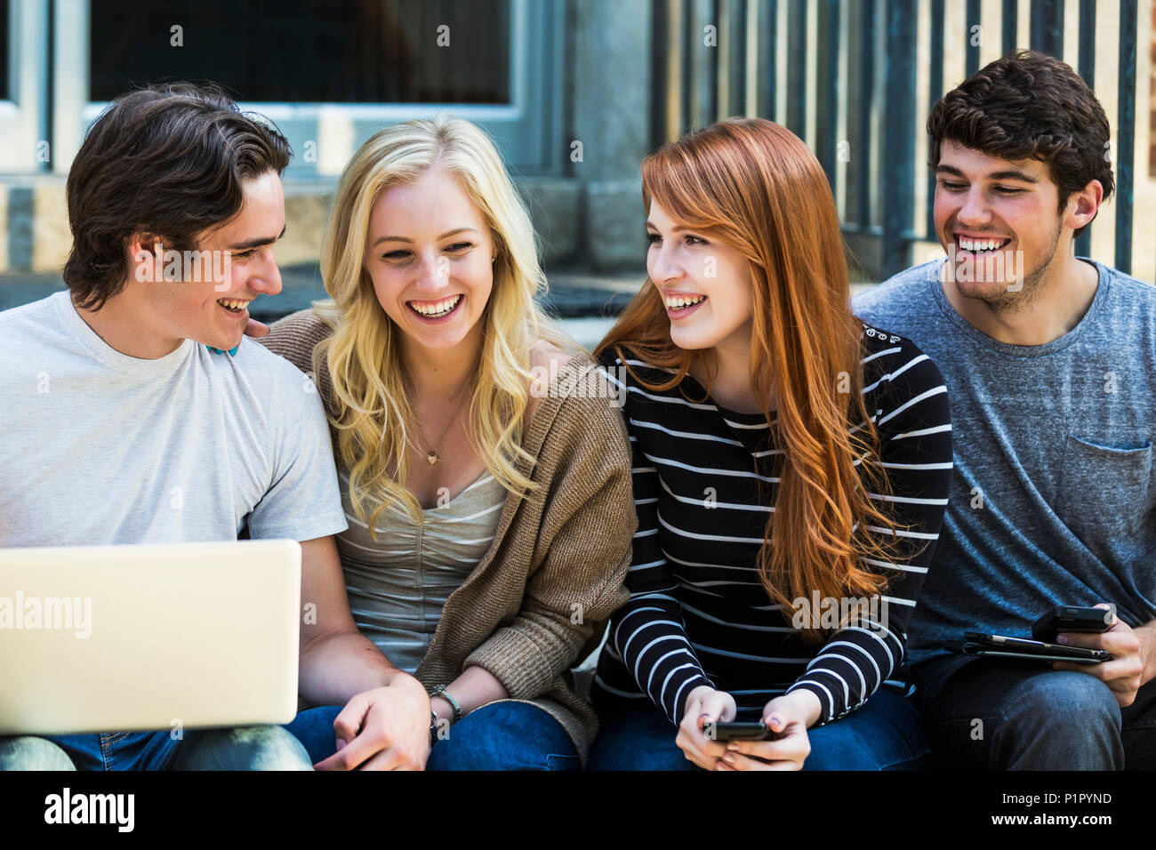 Four students sitting in a row on a step using their technology on the ...