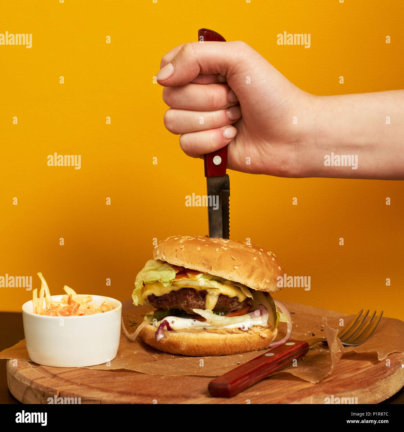 Hand stabbing with knife burger lying with portion of salad and fork on ...