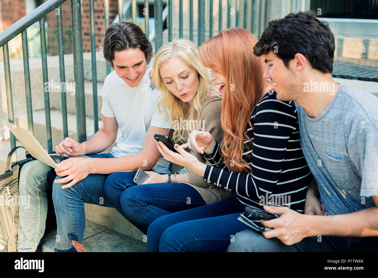 Four students sitting in a row on a step using their technology on the ...