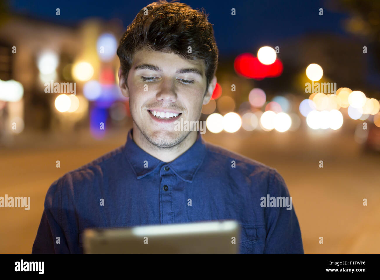 A young man uses a tablet along a street at dusk with the glowing ...