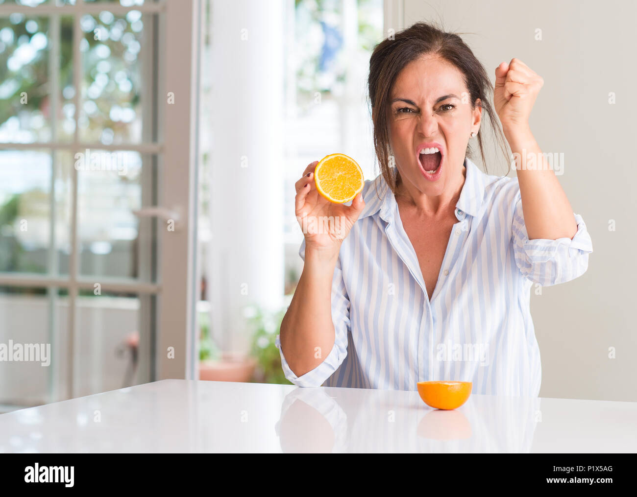 Middle aged woman holding orange fruit annoyed and frustrated shouting ...