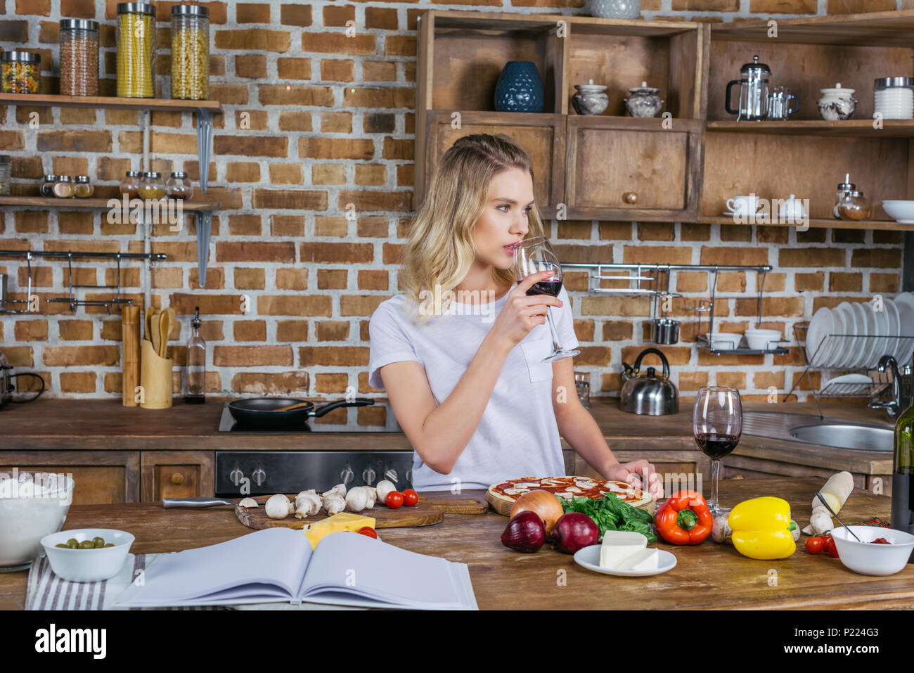 Beautiful young woman drinking wine while cooking pizza at kitchen ...