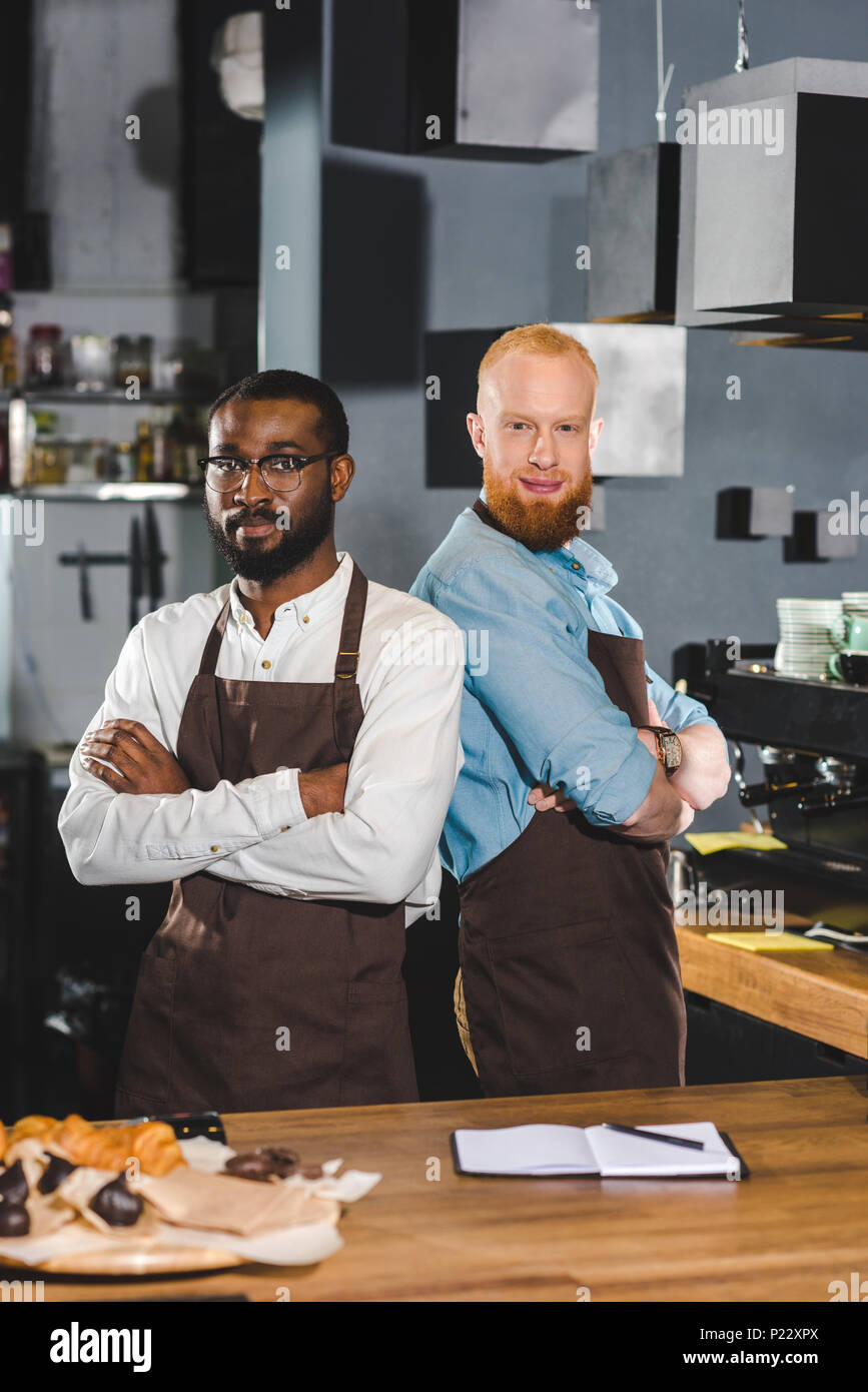 two young multicultural male baristas in aprons standing with crossed ...