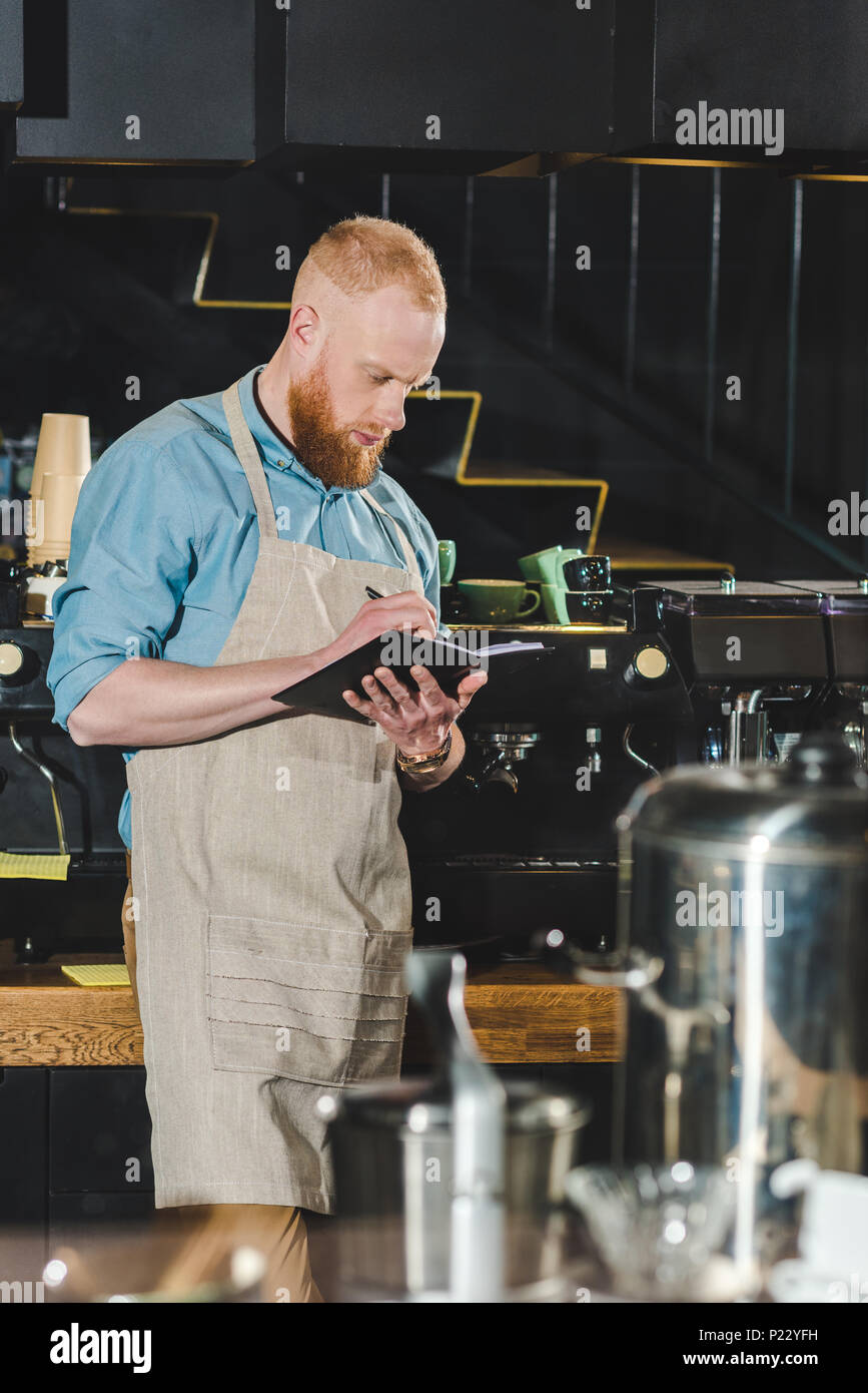 side view of young male barista in apron writing in textbook Stock ...
