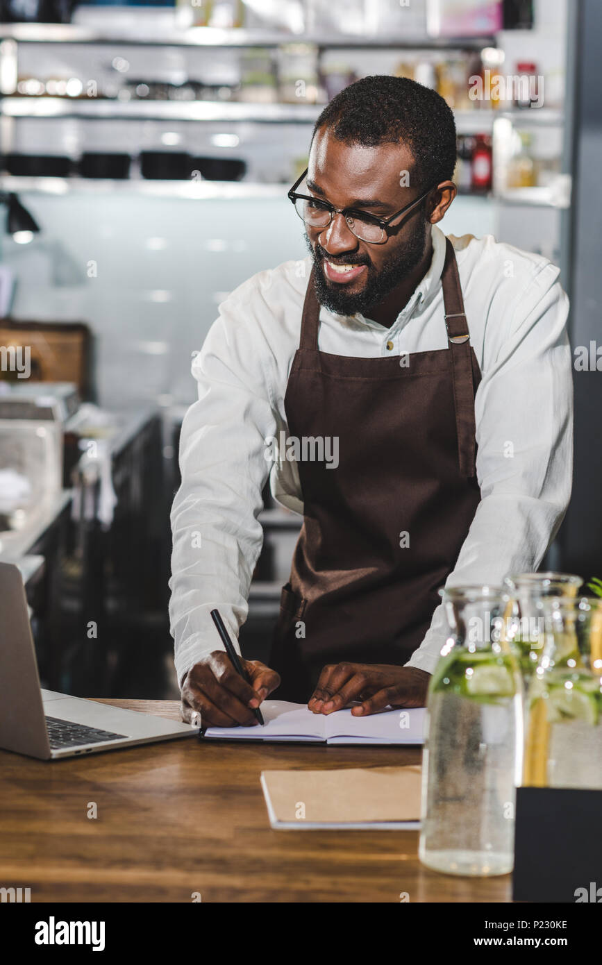 happy young african american barista taking notes and using laptop at ...