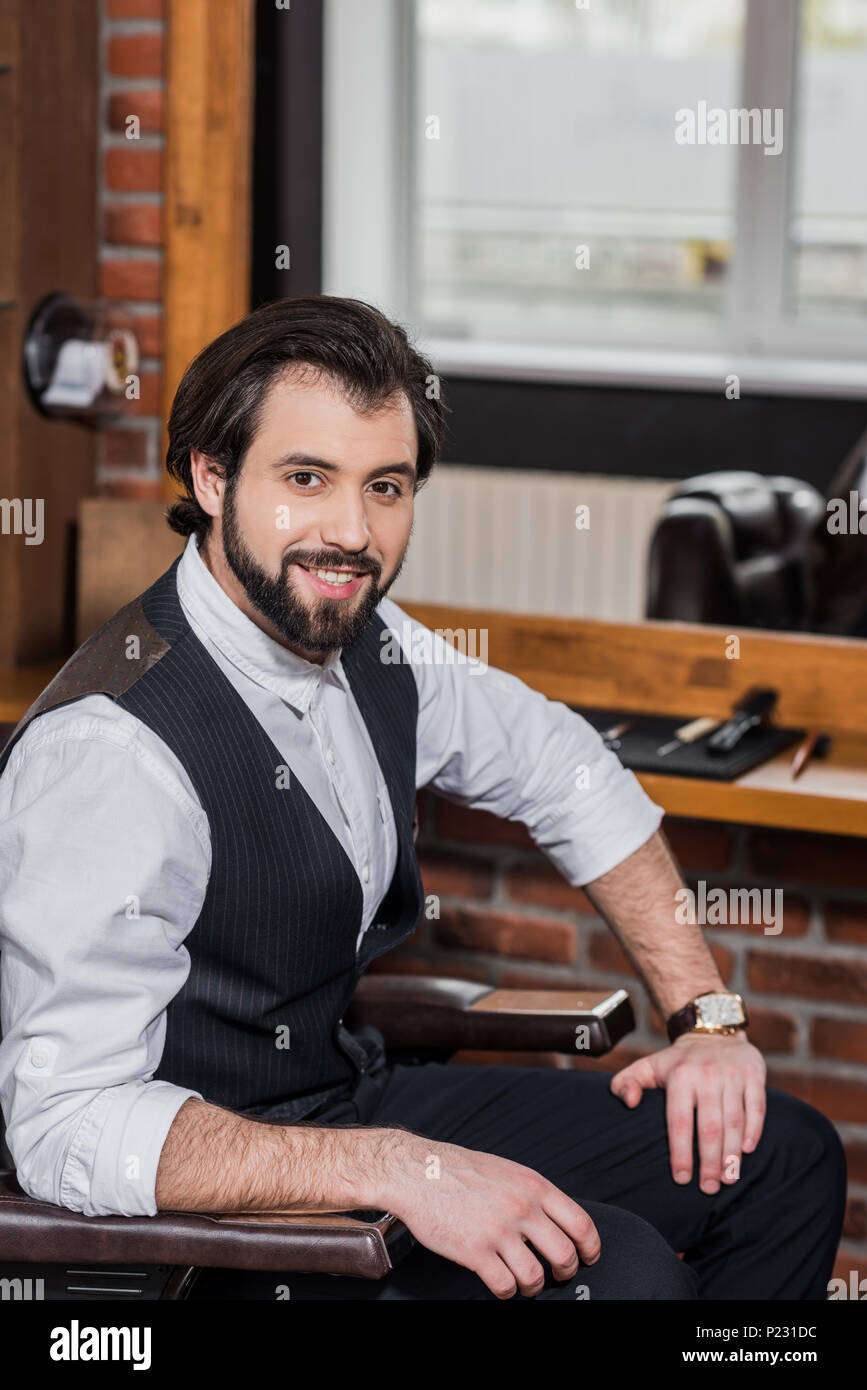 handsome barber sitting on chair at workplace and looking at camera ...