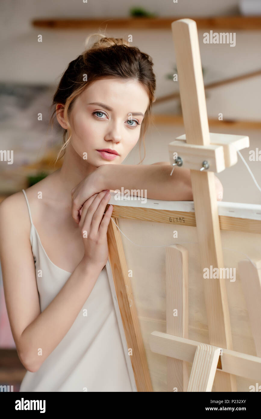 beautiful young woman leaning at picture on easel and looking at camera ...