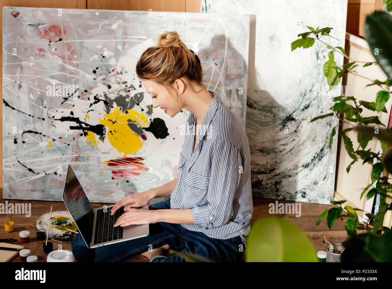 side view of smiling young female painter using laptop in art studio ...