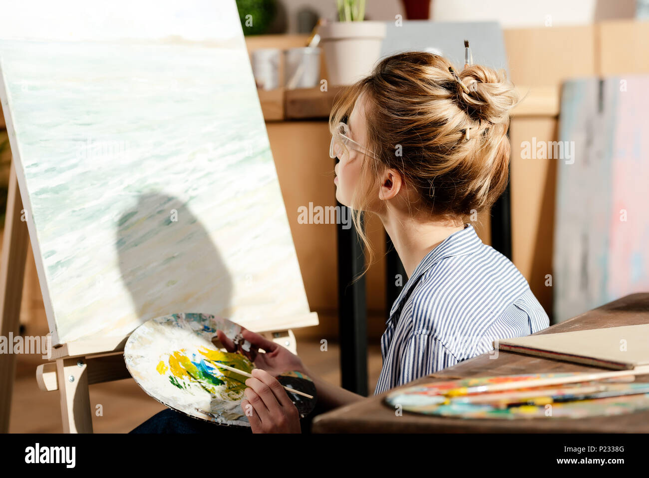 young stylish female artist in eyeglasses using palette and looking at ...