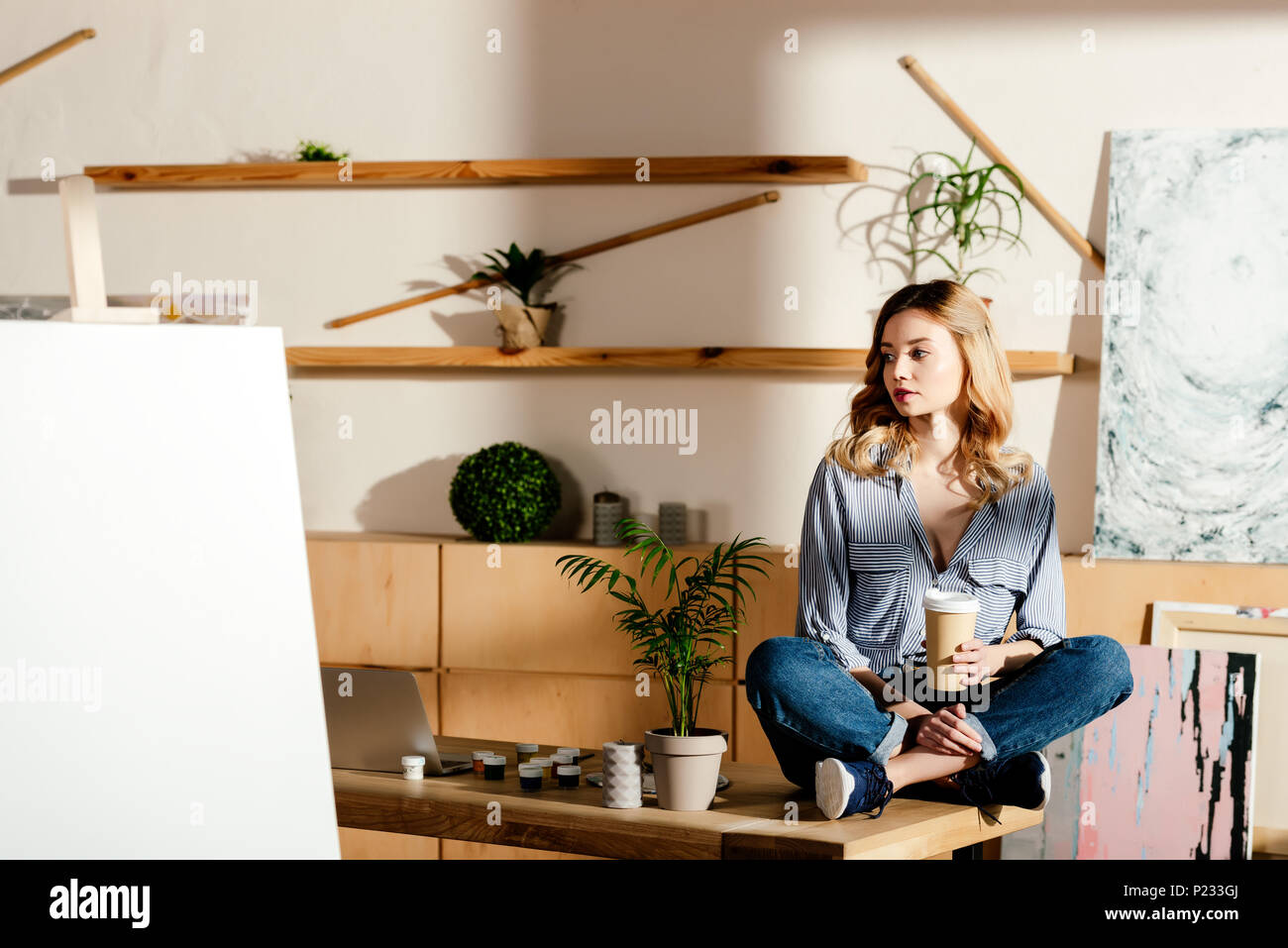 young female artist with paper cup of coffee sitting on table in studio ...