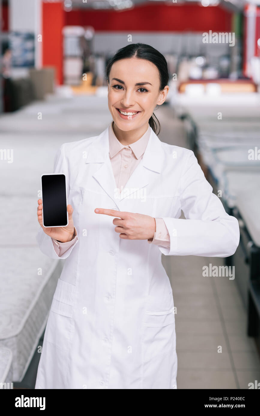 smiling shop assistant in white coat showing smartphone with blank ...