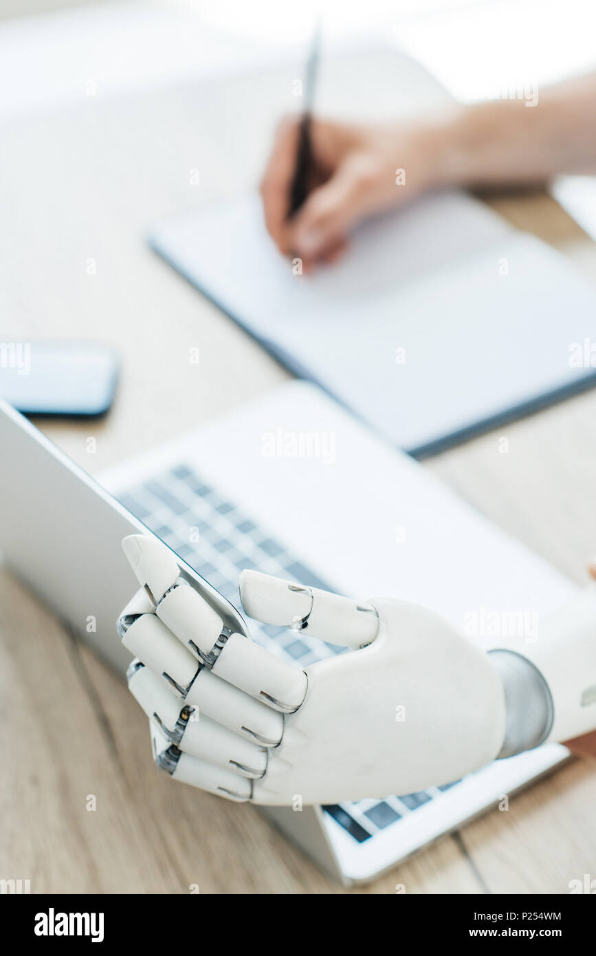 close-up view of robotic arm using laptop and human hand taking notes ...