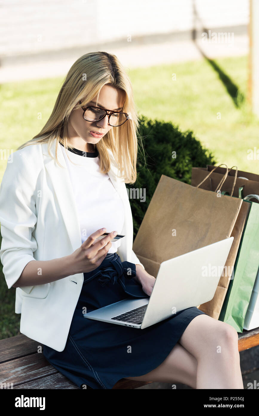 young woman holding credit card and using laptop while sitting on bench ...