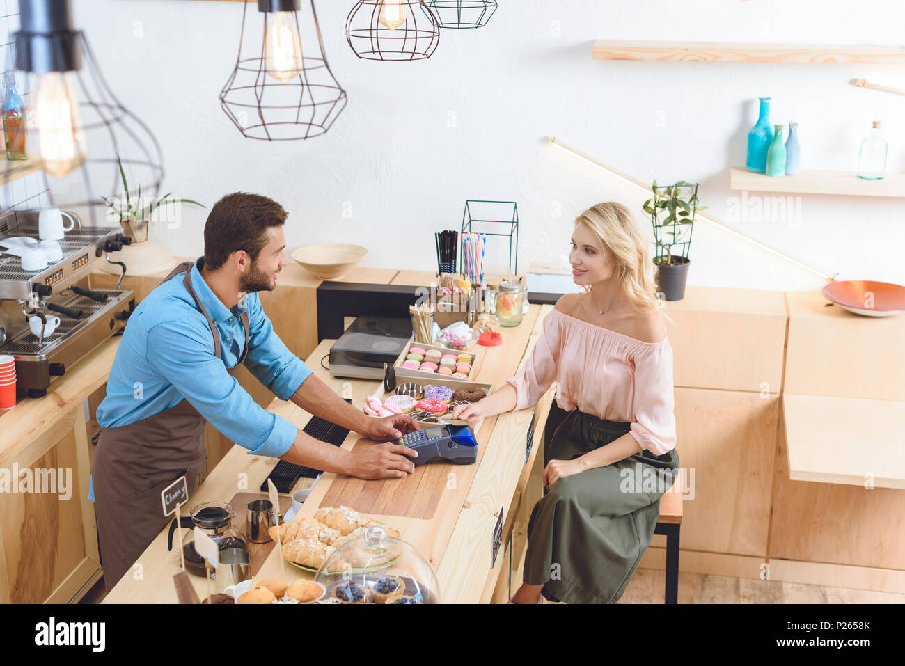 side view of handsome waiter looking at woman paying by credit card in ...