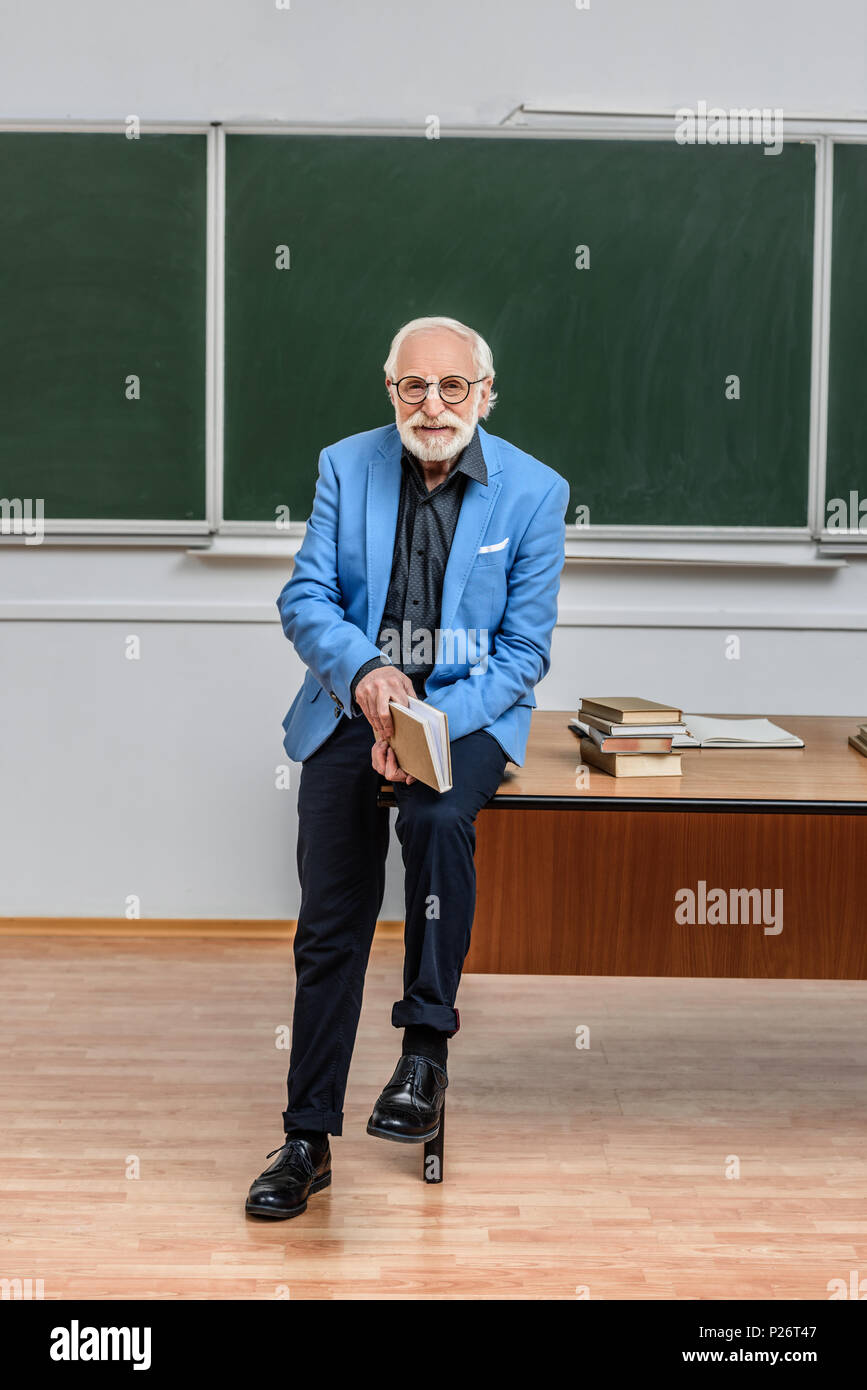 grey hair professor sitting on table in lecture room and holding book ...