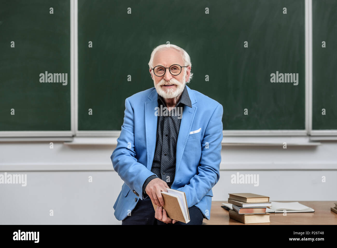 grey hair professor sitting on table in lecture room and holding book ...