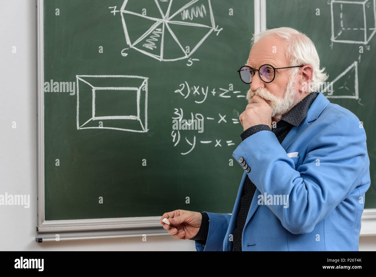 pensive grey hair professor standing at blackboard with piece of chalk ...