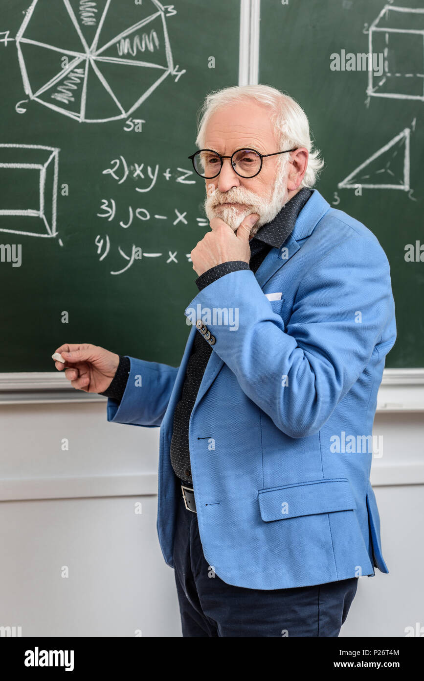 pensive grey hair professor standing at blackboard with piece of chalk ...