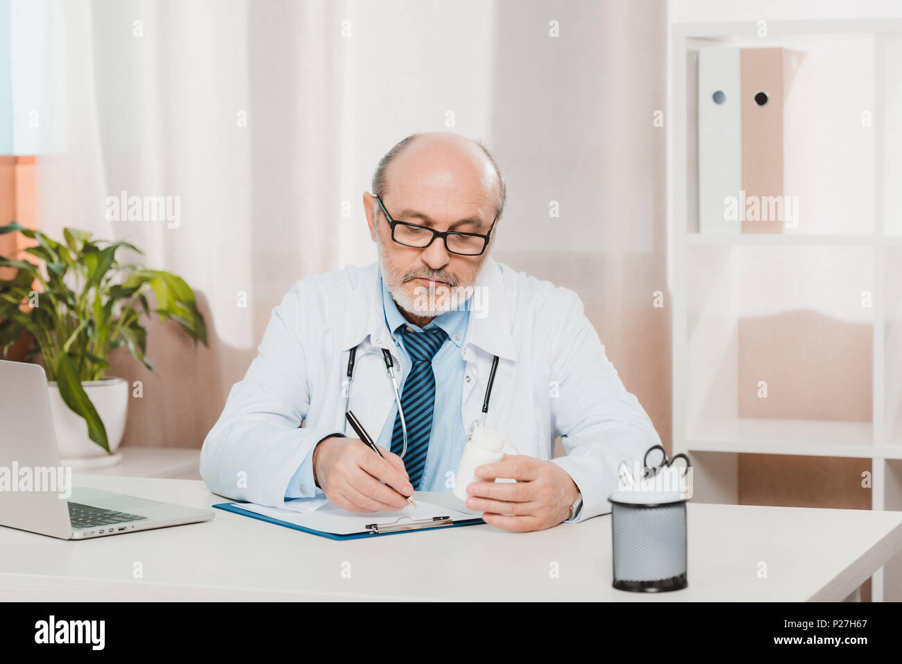 portrait of focused senior doctor making notes on notepad at workplace ...