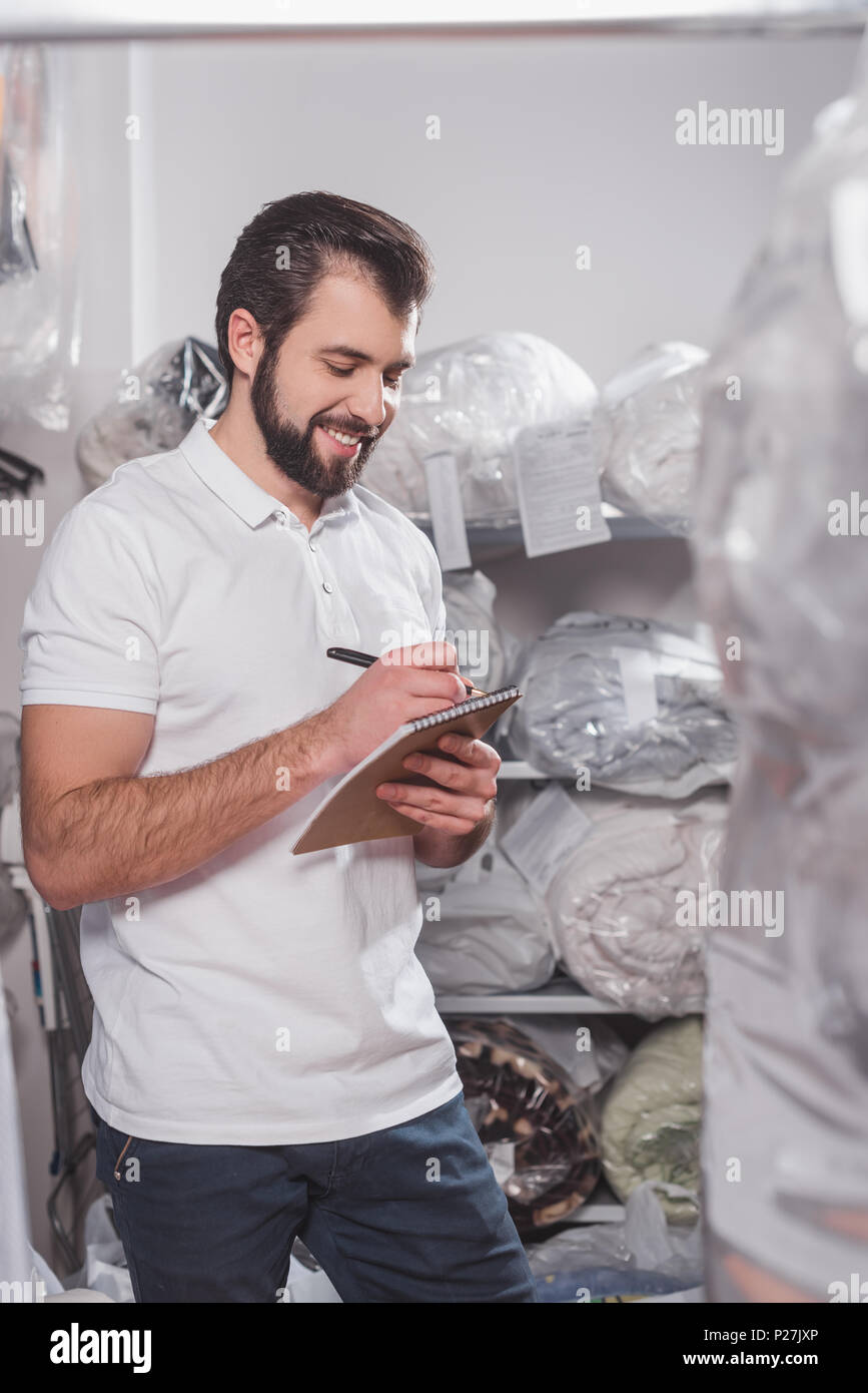 smiling young dry cleaning worker writing in notepad at warehouse Stock ...