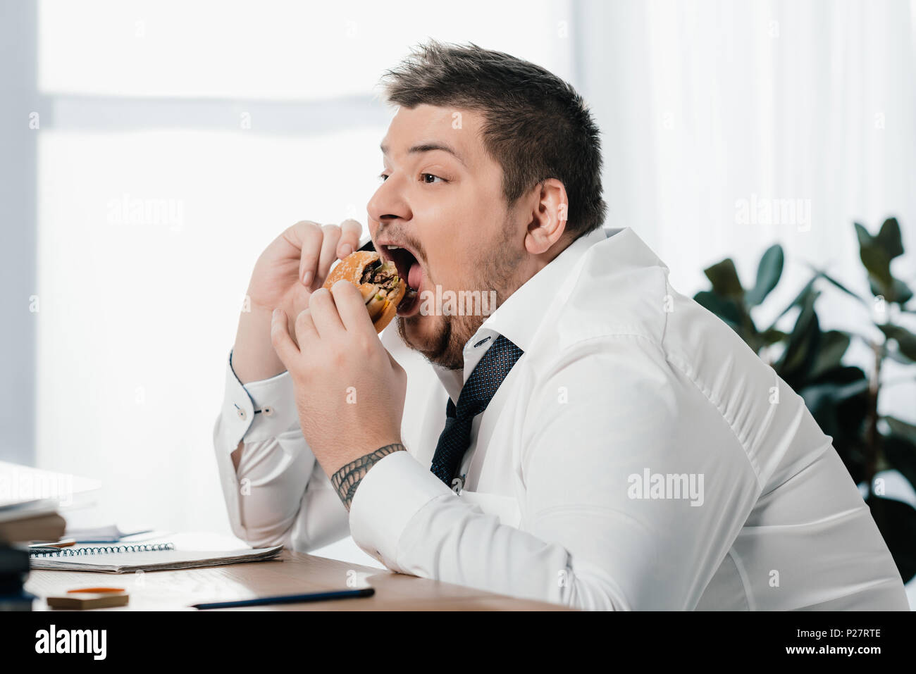 fat businessman talking on smartphone while eating hamburger in office ...