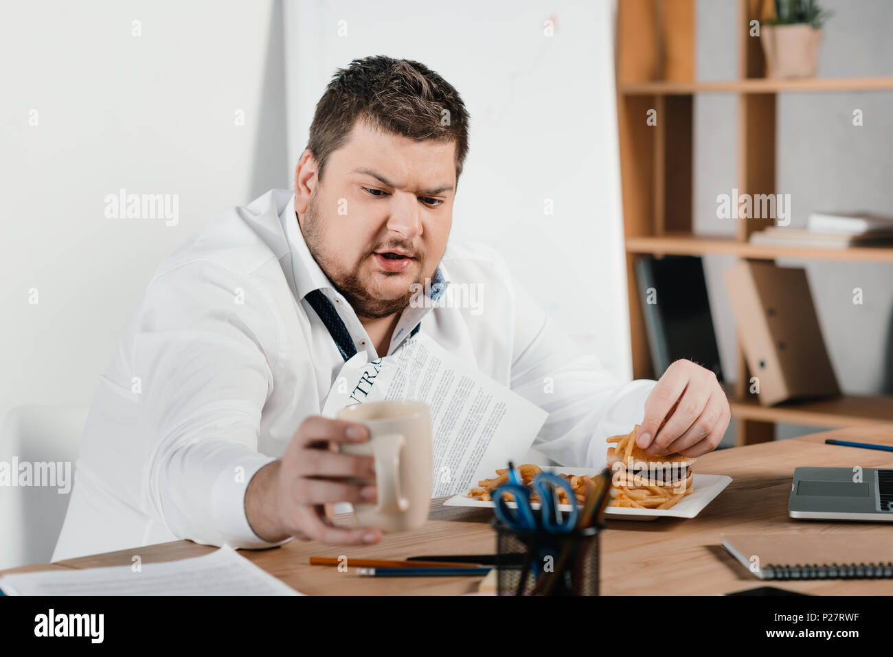 overweight businessman eating fast food and drinking coffee in office ...