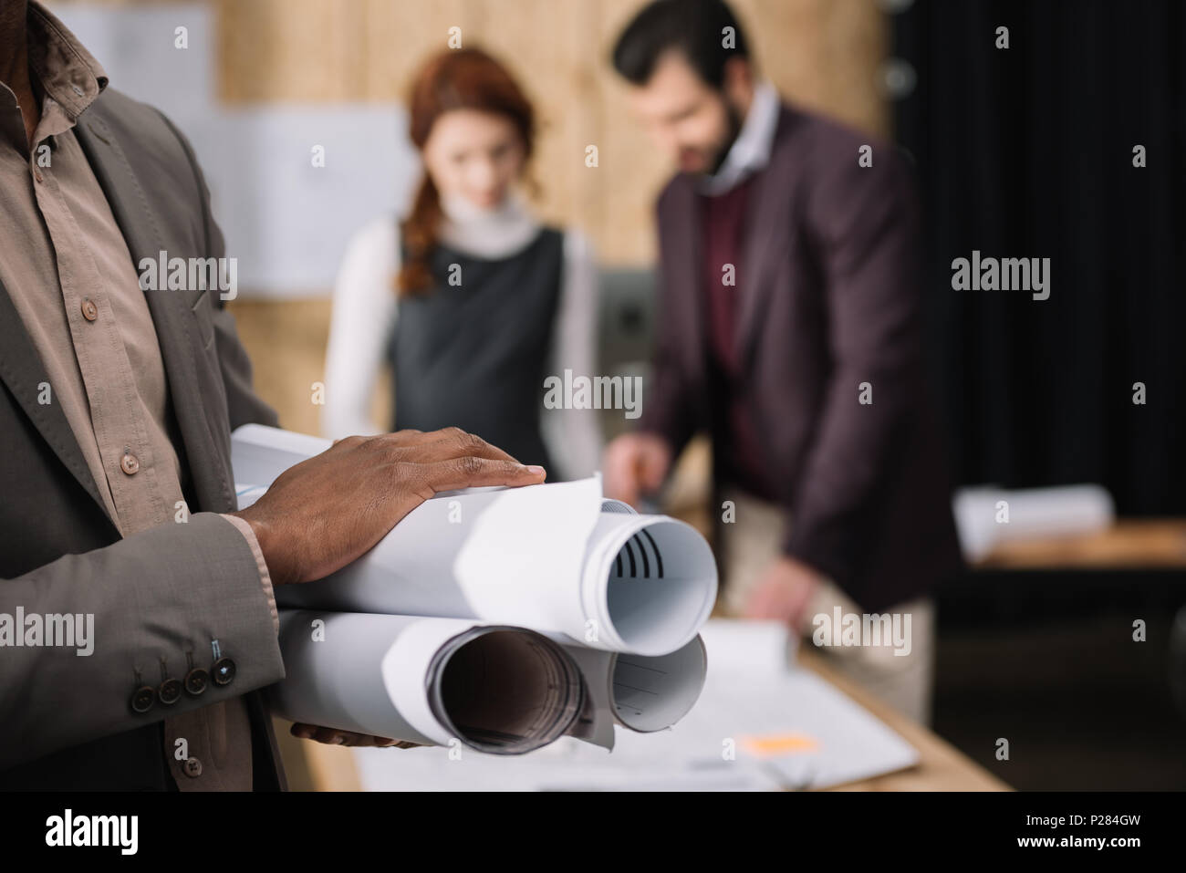 cropped shot of architect holding rolled plans with blurred colleagues ...