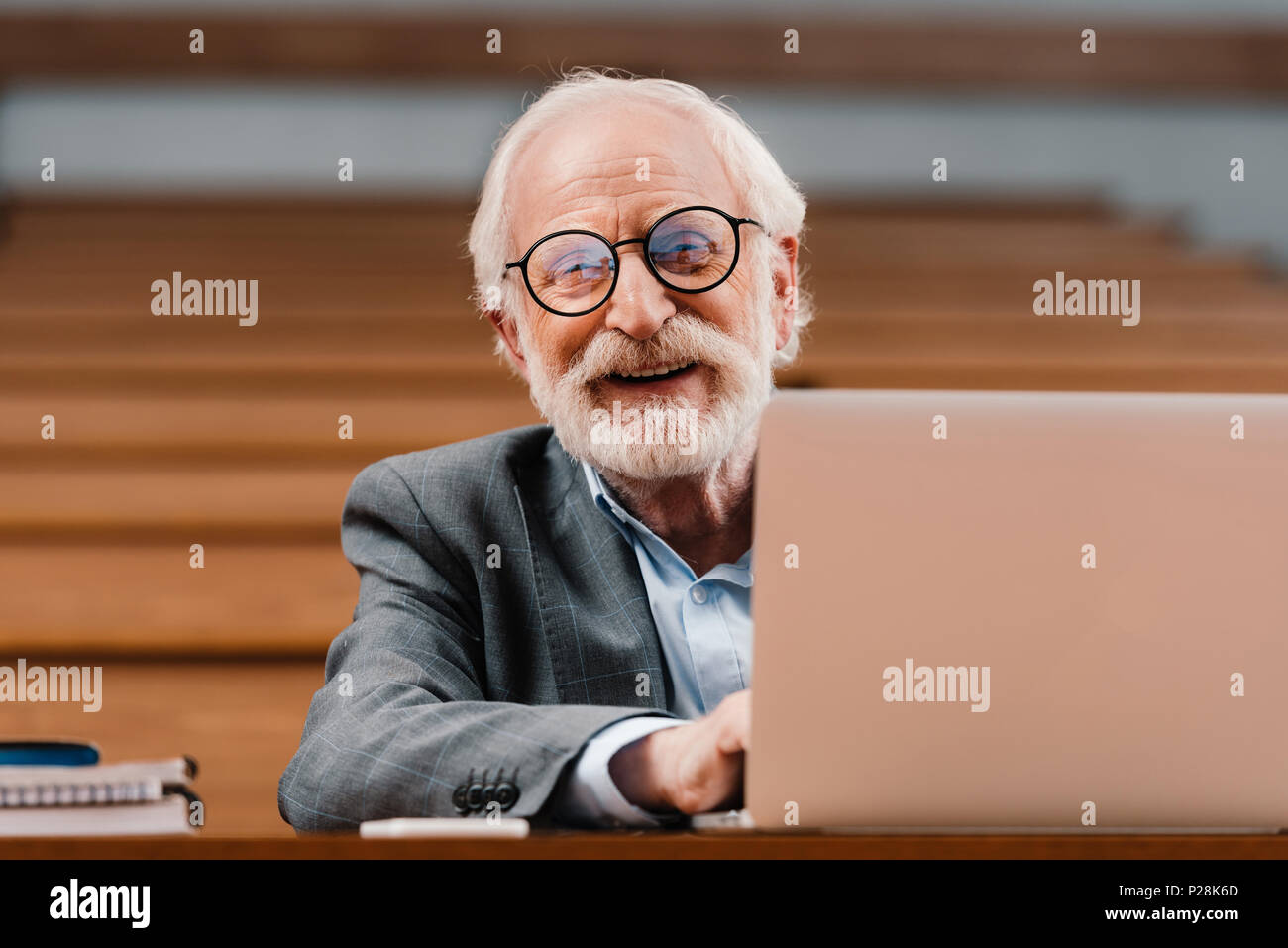 smiling grey hair professor sitting in empty lecture room with laptop ...