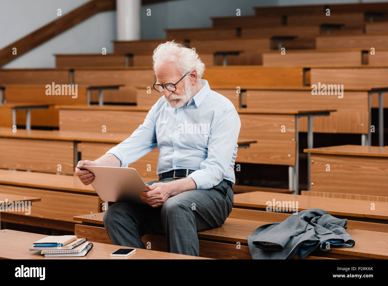 grey hair professor sitting on desk in empty lecture room and using ...
