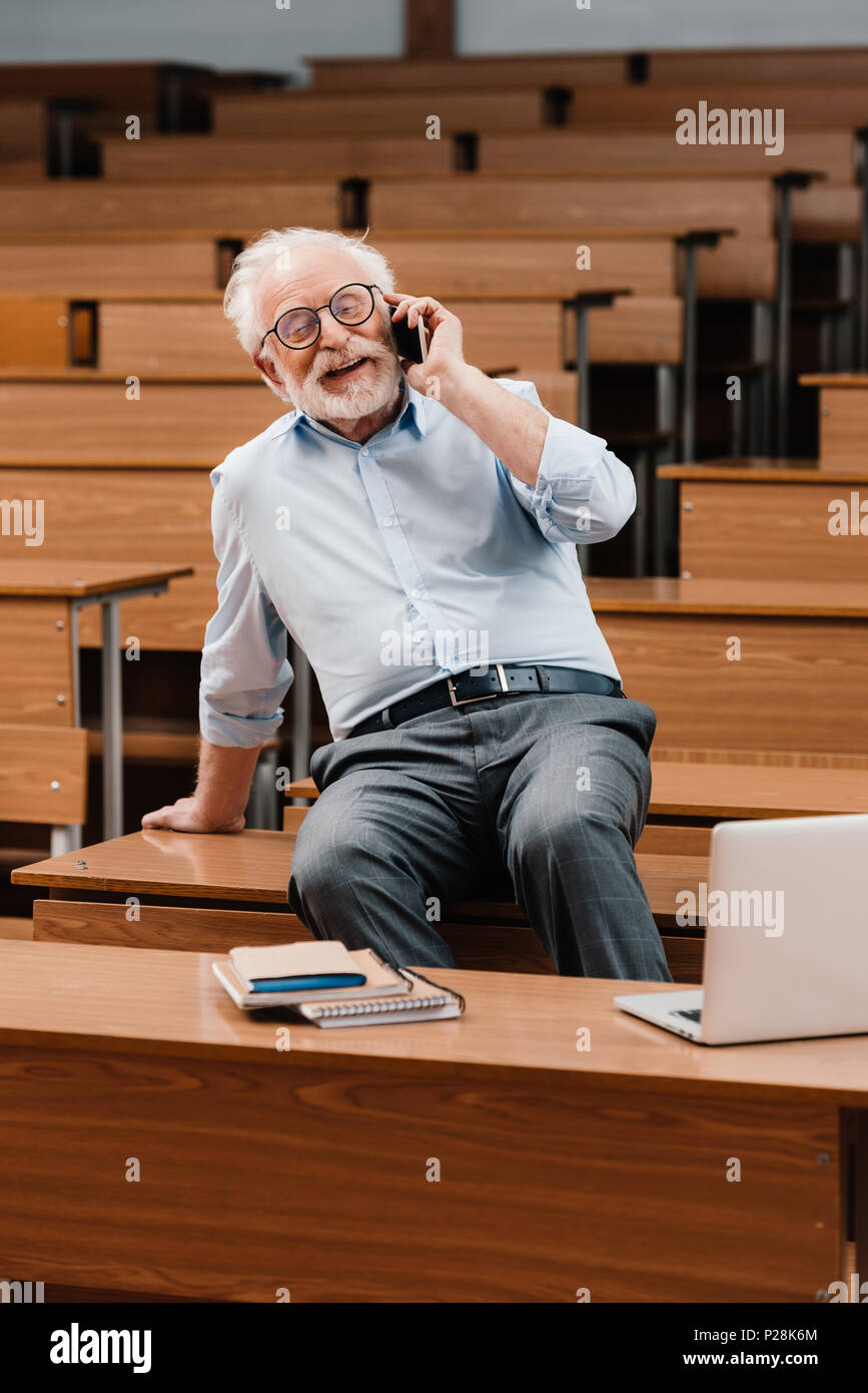 smiling grey hair professor sitting on desk in empty lecture room and ...