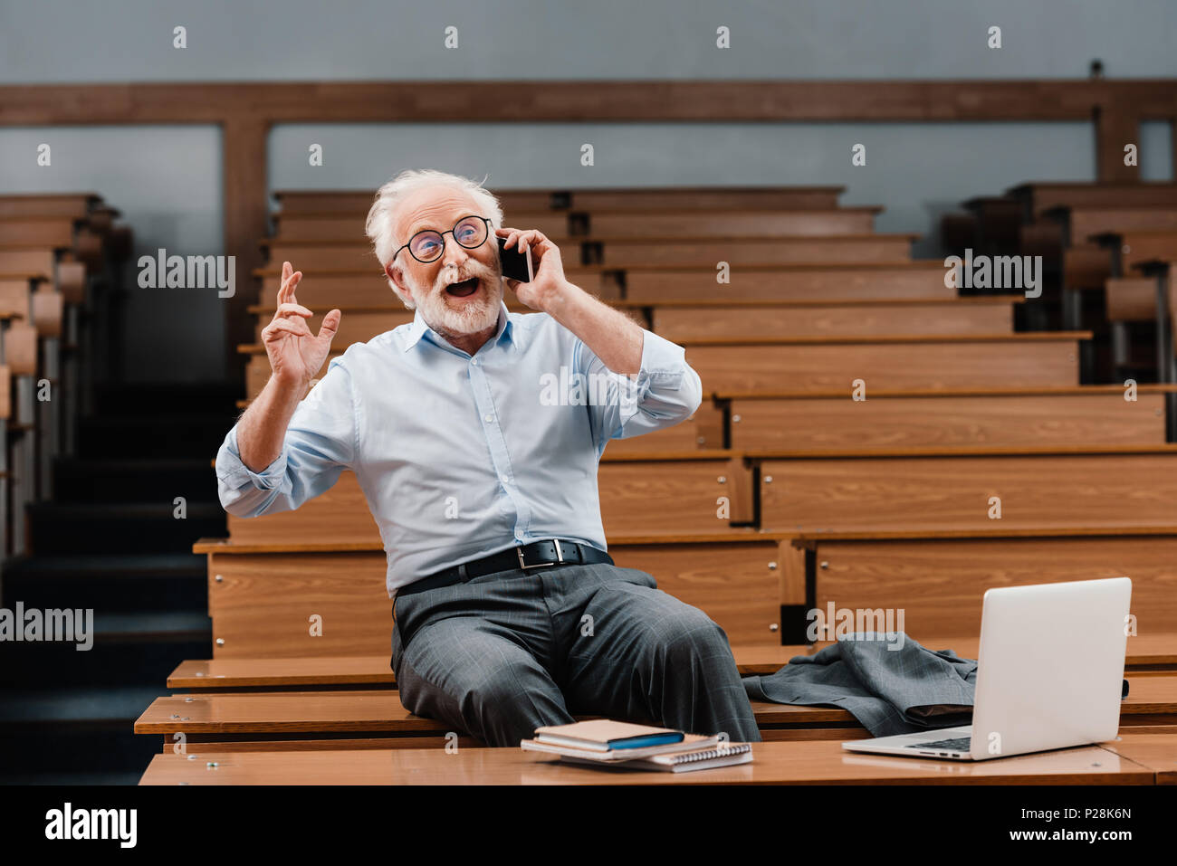 smiling grey hair professor sitting on desk in empty lecture room and ...