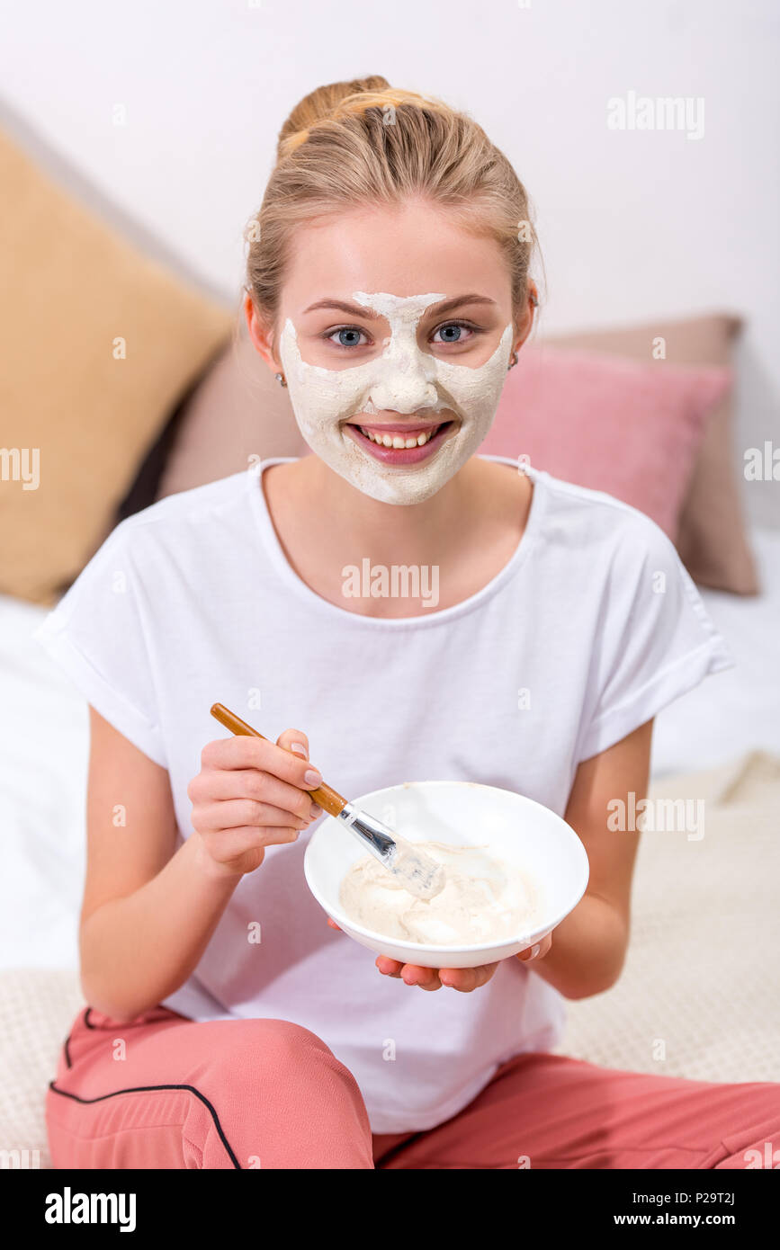 young woman applying clay mask on face at home and looking at camera ...