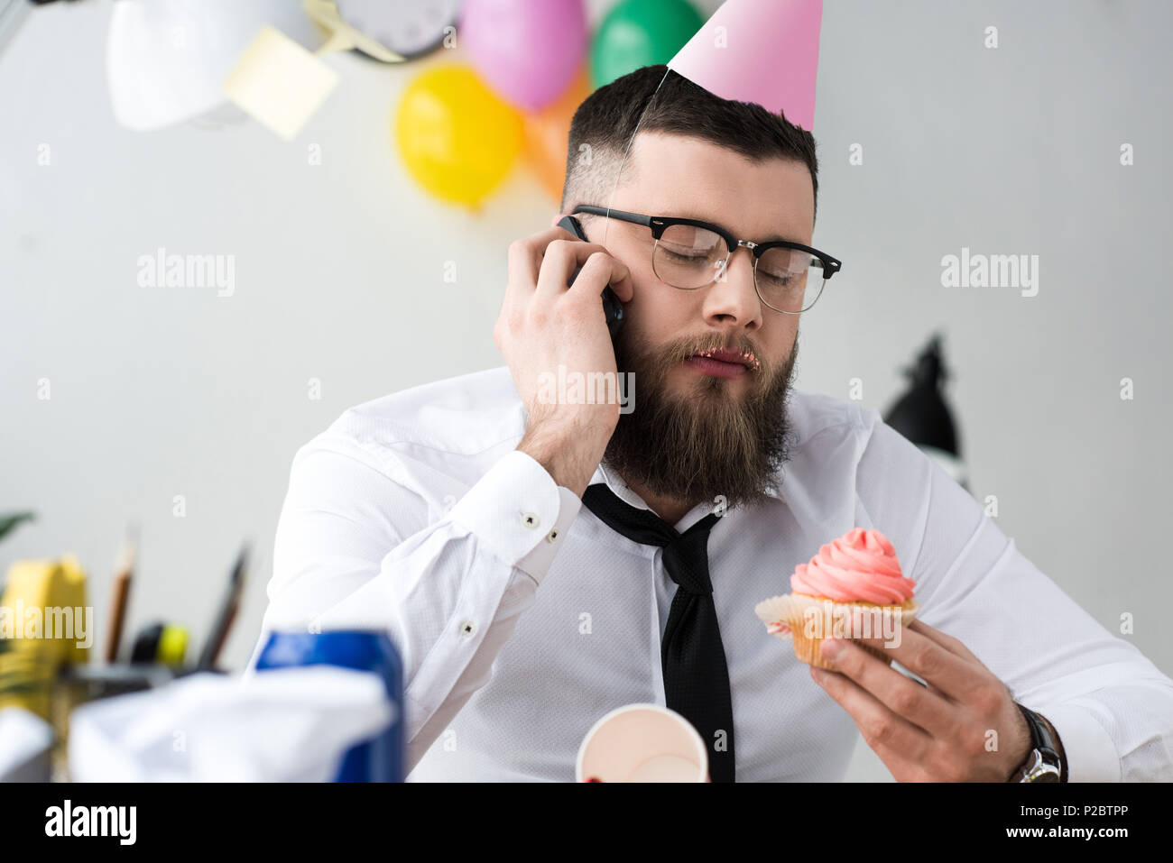portrait of businessman in party cone with birthday cupcake in hand ...